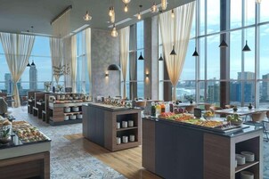 Uma área de refeições de hotel moderna, com grandes janelas, estações de buffet e iluminação contemporânea.
