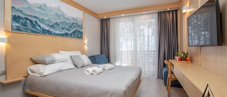 Quarto de hotel moderno com uma cama grande, uma escrivaninha, uma TV e uma pintura de montanhas na parede.