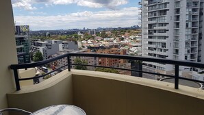 Uma varanda com guarda-corpo de metal, uma mesa redonda e vista para a paisagem urbana, incluindo diversos edifícios e um céu claro.