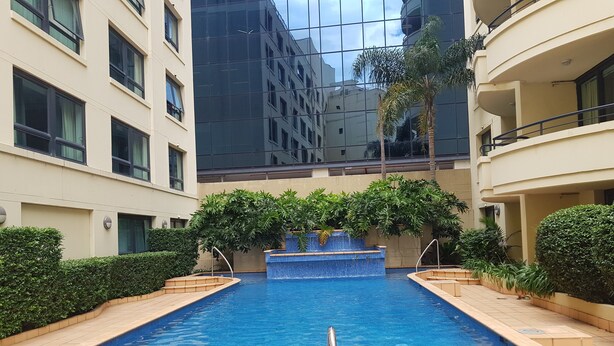 Piscina com borda azulejada, cercada por vegetação e um edifício moderno com vários andares e varandas.