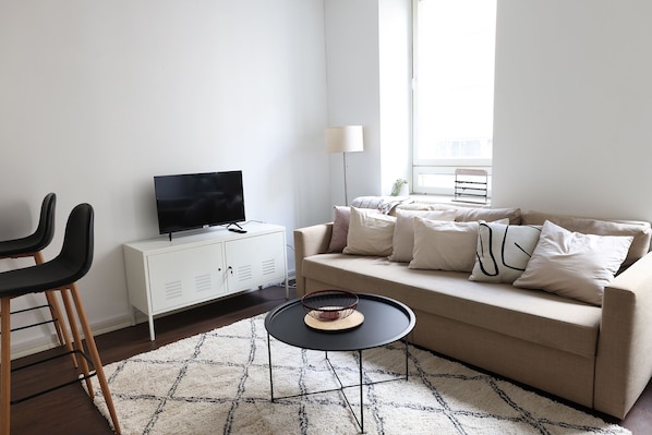 Uma sala de estar moderna com um sofá, uma televisão sobre um móvel branco, uma mesa de centro e um abajur.