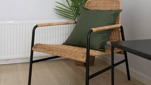 Uma cadeira de vime com almofada, uma mesa pequena e uma planta verde alta em um quarto com paredes brancas e piso de madeira.