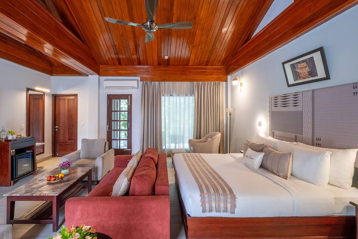 Quarto de hotel com teto de madeira, cama com roupa de cama branca, sofá, cadeira, mesinha com uma fruteira e um quadro na parede.