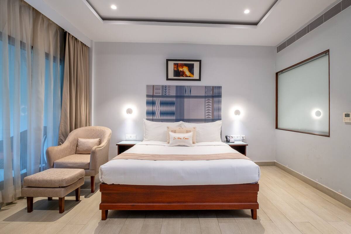 Quarto de hotel moderno com uma cama de madeira, uma poltrona bege e uma janela grande com cortinas leves.