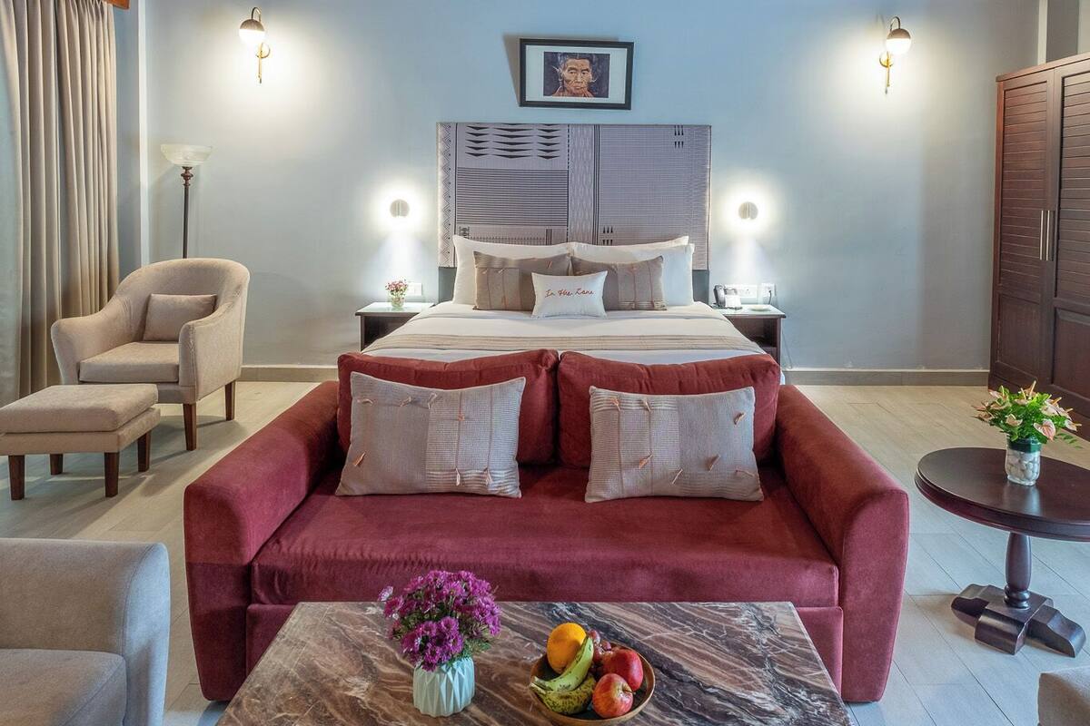 Um quarto espaçoso com uma cama grande, um sofá vermelho, uma mesa de centro de mármore com frutas e um ventilador de teto.