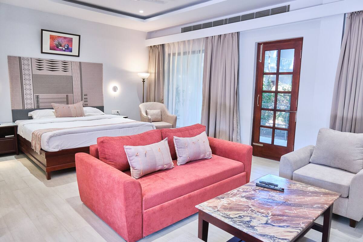 Quarto de hotel moderno com uma cama grande, um sofá vermelho, uma poltrona bege e uma mesa de centro de mármore.