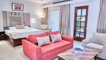 Quarto de hotel moderno com uma cama grande, um sofá vermelho, uma poltrona bege e uma mesa de centro de mármore.