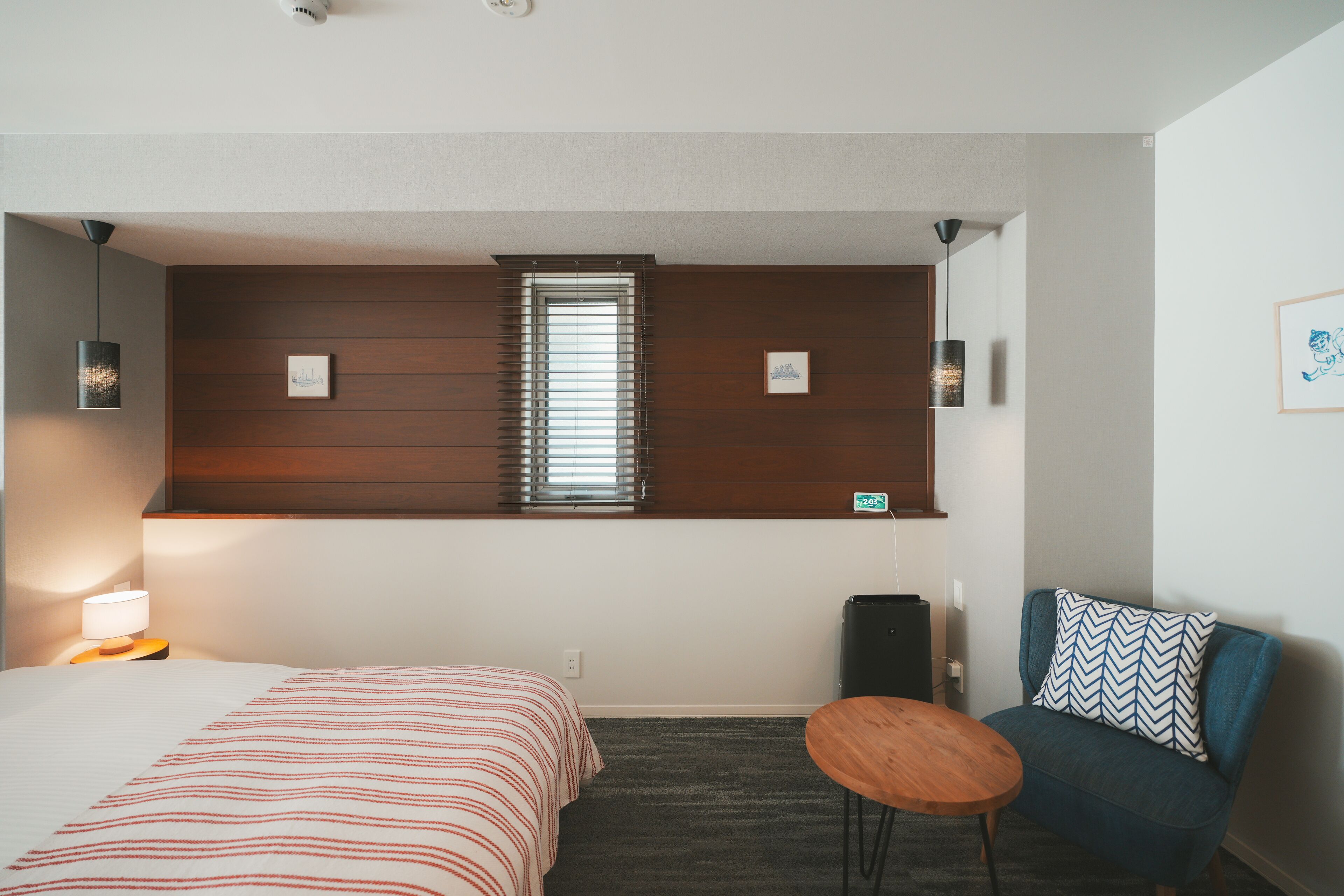 Quarto de hotel com cama, uma mesa pequena, uma cadeira, janela com persianas e um alto-falante fixado na parede.