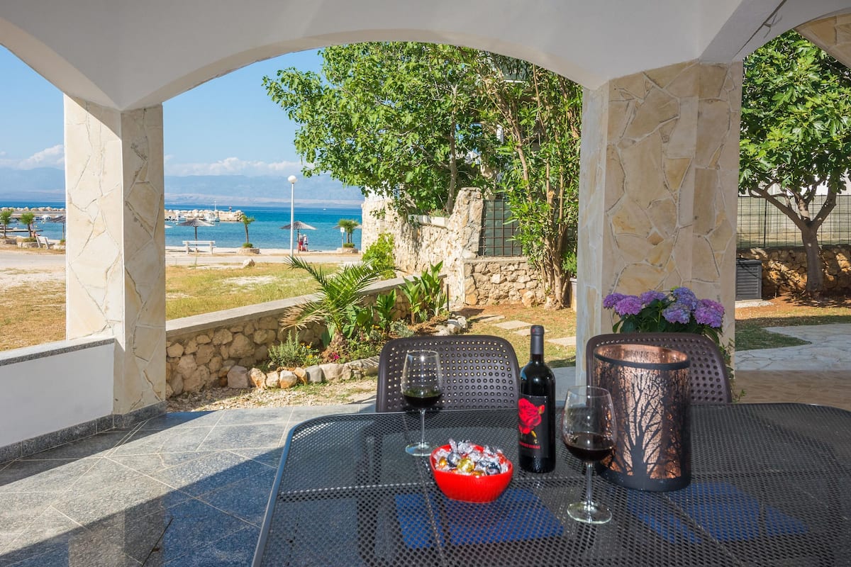 Mesa com garrafa de vinho, um copo e uma tigela de petiscos, com vista panorâmica para o mar e uma parede de pedra.