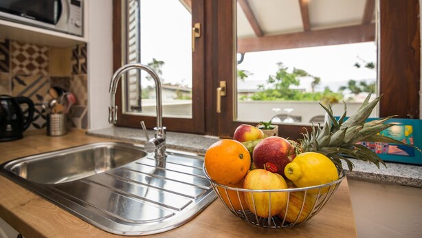 Uma cozinha com pia de aço inoxidável, uma fruteira com maçãs, laranjas e uma abacaxi, e um micro-ondas sobre a pia.