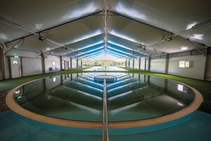 Uma grande piscina interior com uma plataforma de mergulho central, rodeada por espreguiçadeiras e uma área coberta com vista para o exterior.