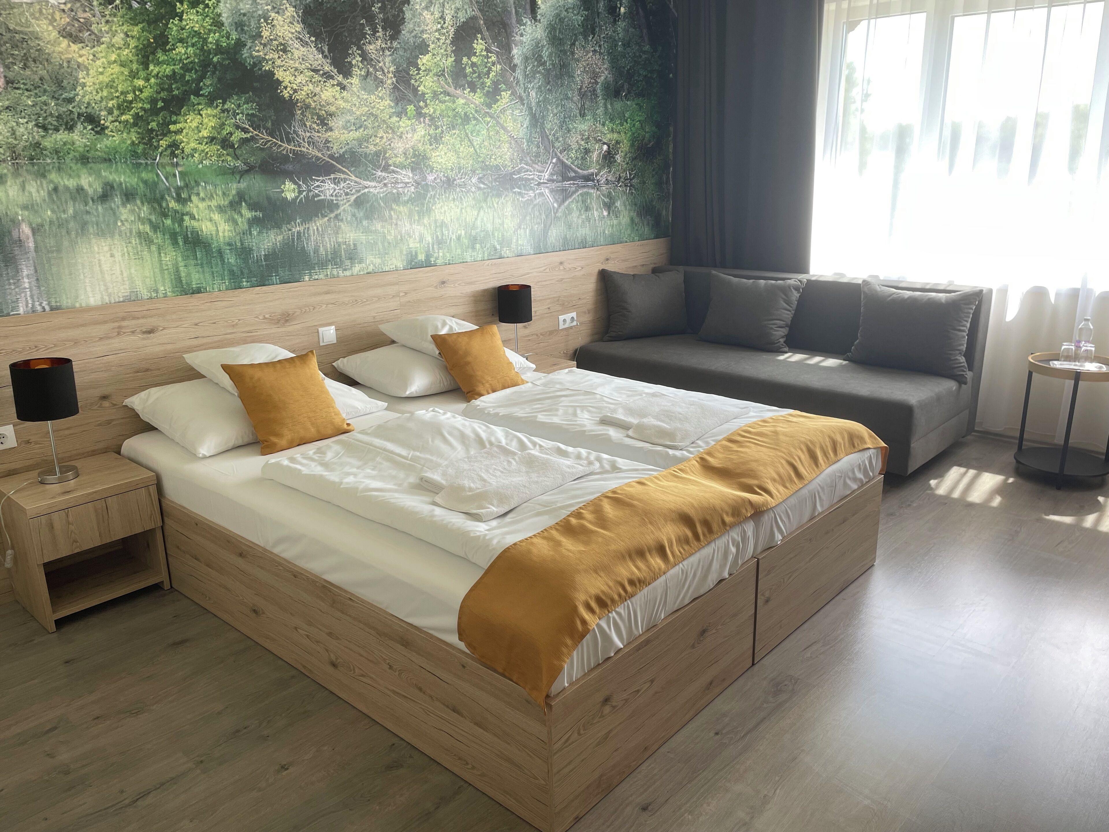 Quarto de hotel moderno com uma cama grande, um sofá, uma mesa de cabeceira, um abajur e uma janela ampla.