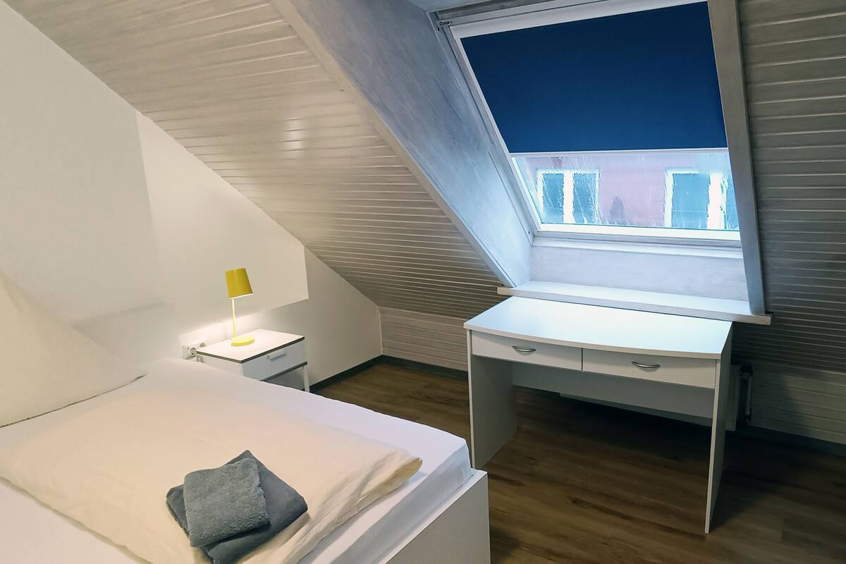 Quarto com teto inclinado, claraboia, cama, escrivaninha e mesa de cabeceira.