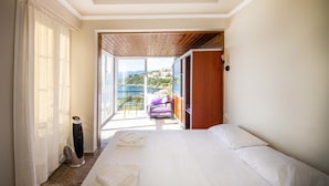 Quarto com cama, ventilador, cadeira e vista para o mar através de uma porta de vidro.