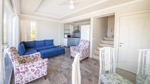 Uma sala de estar ampla, com um sofá azul, poltronas florais, uma mesa de centro de vidro e um ventilador de teto.