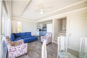 Uma sala de estar ampla, com um sofá azul, poltronas florais, uma mesa de centro de vidro e um ventilador de teto.
