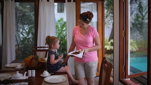 Mulher de camisa e shorts rosa atende uma criança em uma mesa, com uma tigela de frutas, pratos e copos.