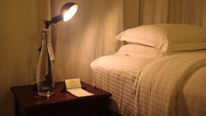 Cama bem arrumada com lençóis brancos, abajur, mesinha com luminária e um aviso sobre a mesa.