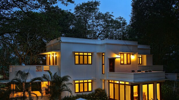 Uma casa moderna de dois andares com janelas amplas e uma varanda, rodeada por vegetação exuberante.