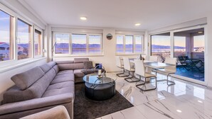 Uma sala de estar moderna com um sofá seccional, uma mesa de centro de vidro e uma área de jantar com mesa e cadeiras brancas. Grandes janelas oferecem vista para as montanhas.