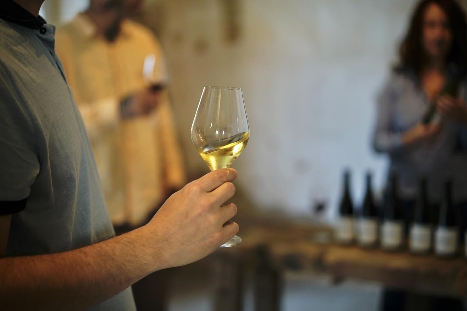 Uma pessoa segurando uma taça de vinho em um bar, com garrafas ao fundo.