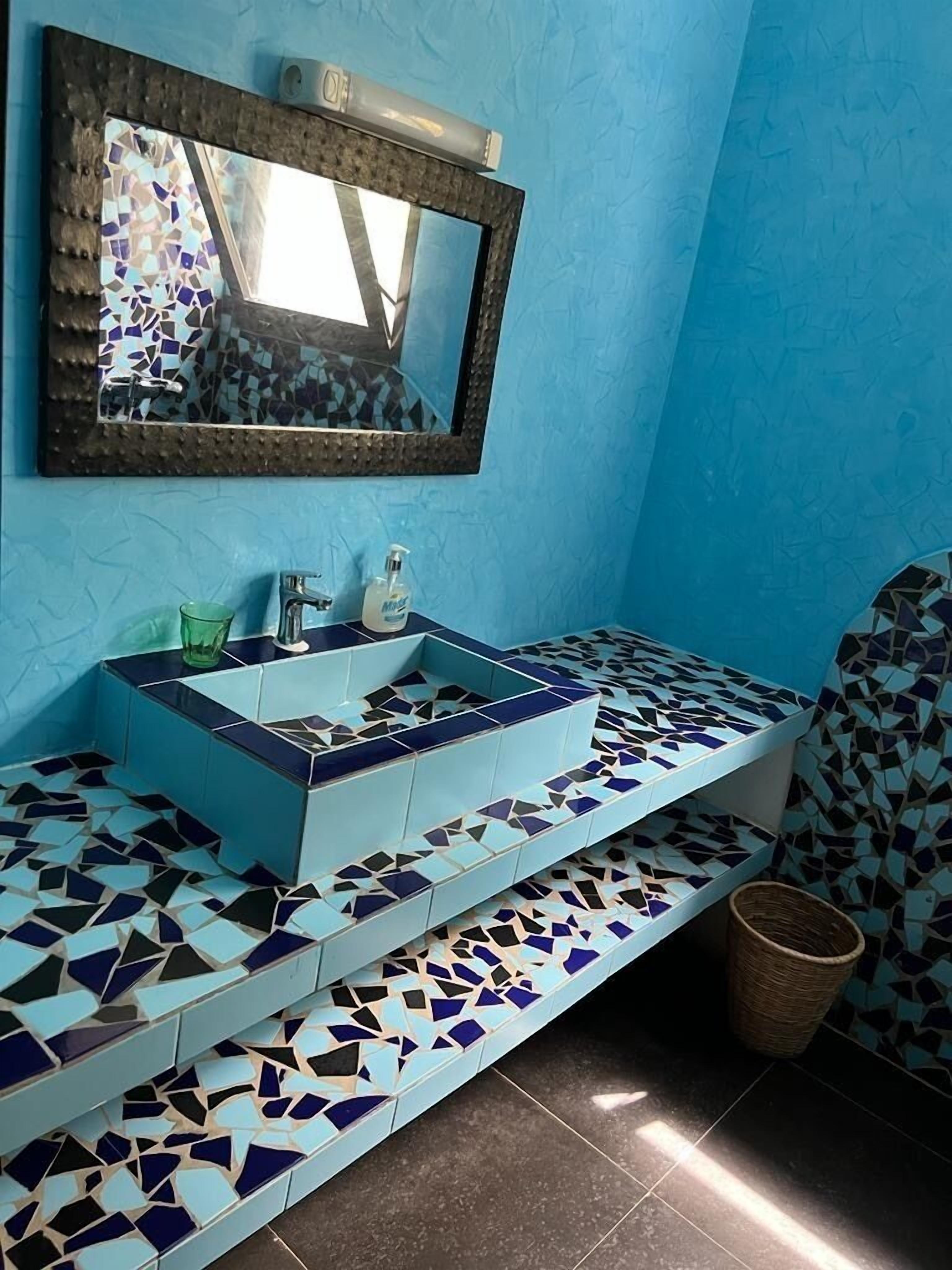Um banheiro com piso azul ladrilhado, pia com espelho acima e cesta de vime no chão.