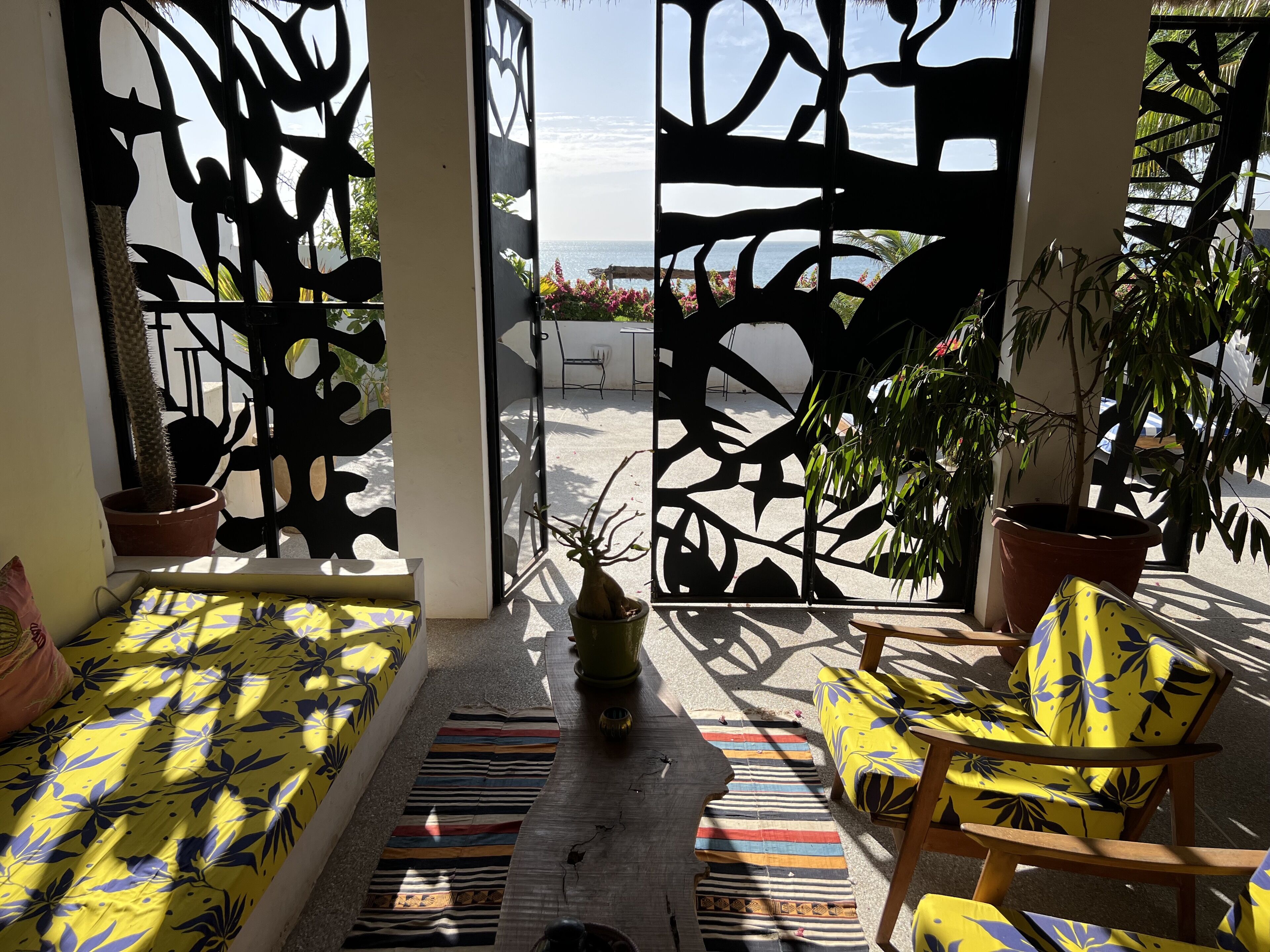 Pátio iluminado pelo sol com vista para o mar, com um sofá estampado, uma cadeira e plantas em vasos.