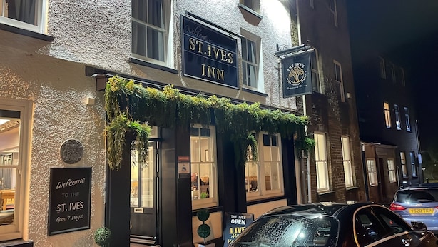Exterior do St. Ives Inn com um letreiro dando as boas-vindas aos visitantes, um carro estacionado na frente e um aviso indicando que a hospedaria está aberta 7 dias por semana.