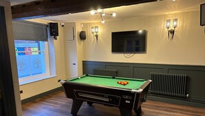 Uma mesa de bilhar com superfície de feltro verde, uma bola de cue branca e um conjunto de bolas vermelhas e amarelas. Há uma televisão fixada na parede acima da mesa.