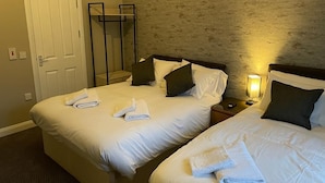 Quarto de hotel com duas camas, cada uma com toalhas dobradas, uma mesa de cabeceira com abajur e uma porta à esquerda.