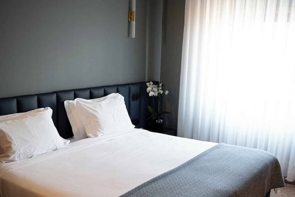 Uma cama bem arrumada com lençóis brancos e um cobertor cinza, um espaldar escuro e uma janela com cortinas leves.
