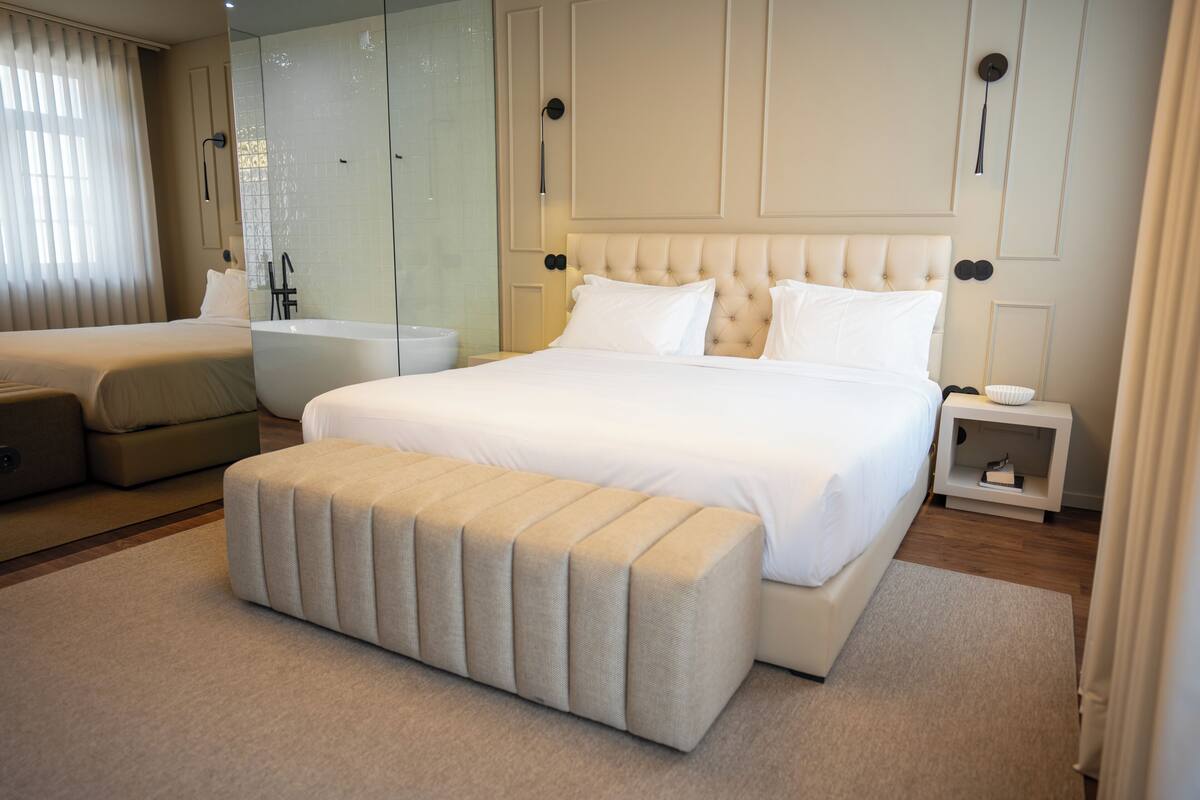 Quarto de hotel com cama grande, sofá separado, chuveiro em cabine de vidro e pequena mesa de cabeceira.