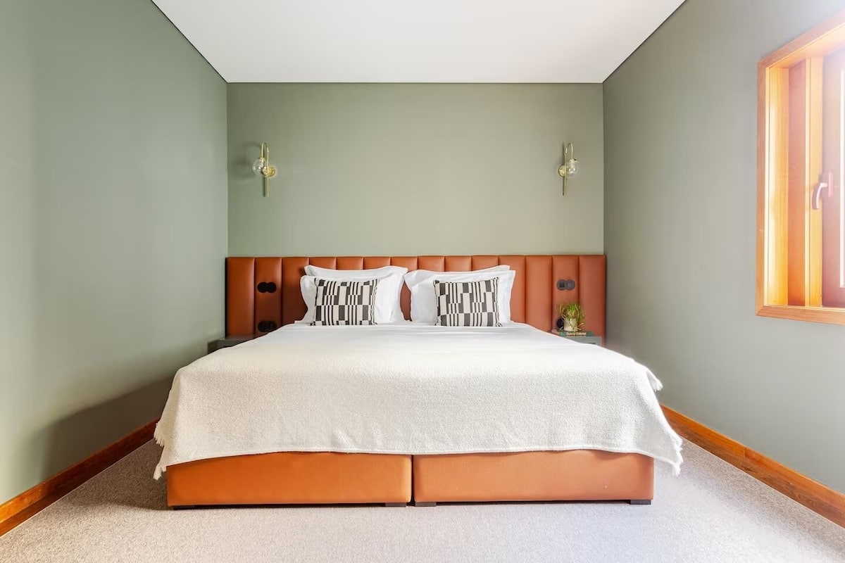 Cama bem arrumada com colcha branca e duas almofadas estampadas, cabeceira estofada marrom e pequena mesa de cabeceira com uma planta.