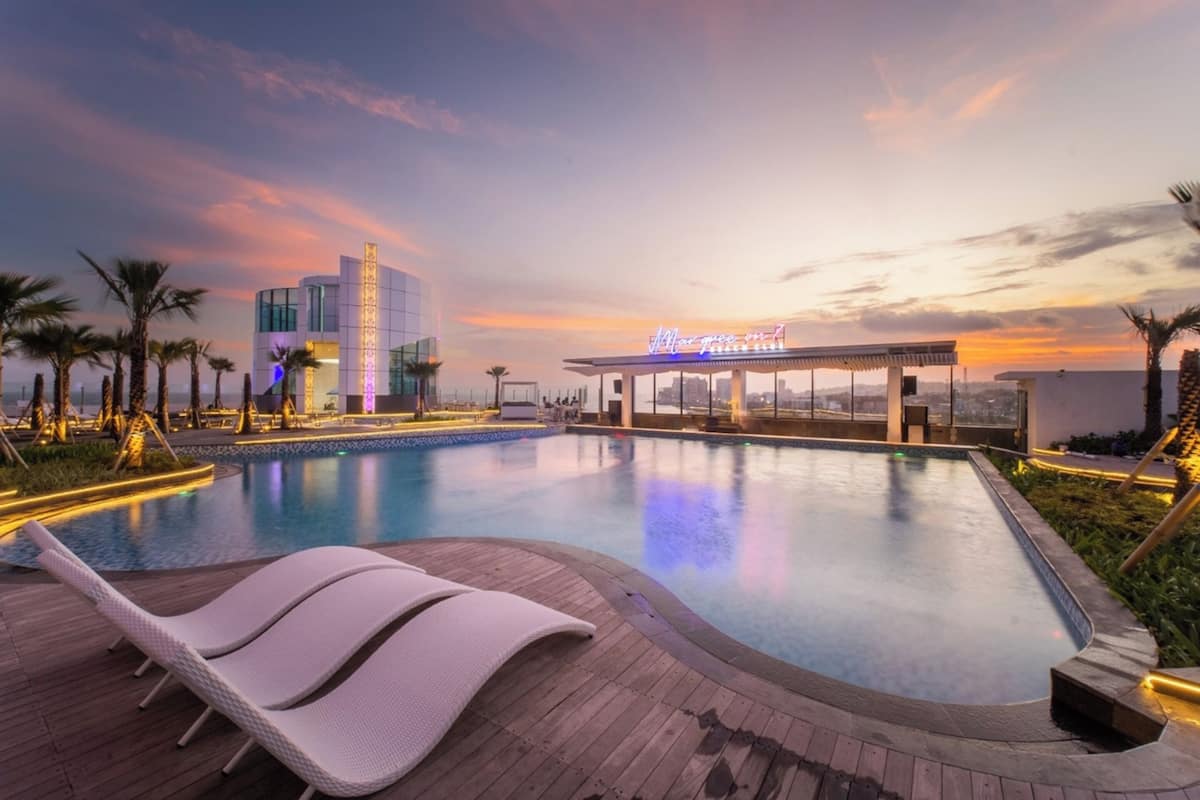 Um hotel moderno com piscina, espreguiçadeiras e vista para o horizonte da cidade ao pôr do sol.