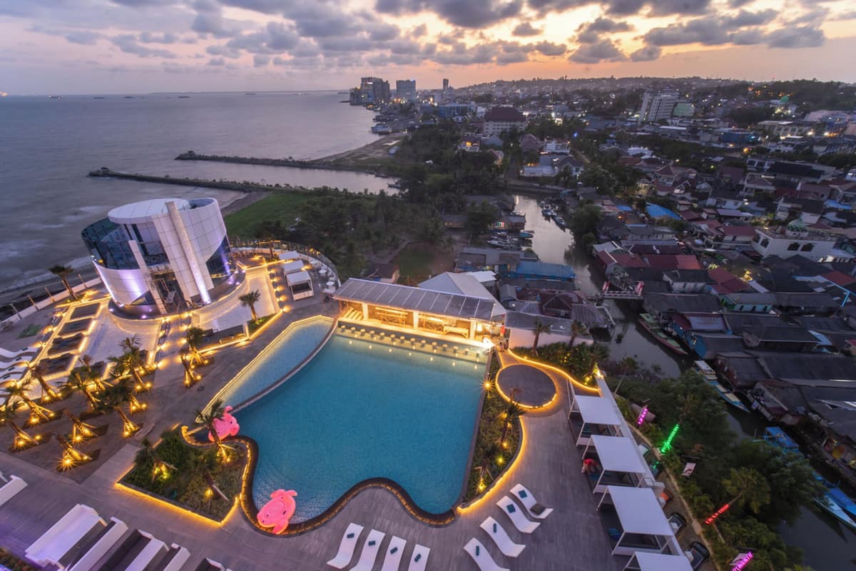 Vista aérea de um resort com uma grande piscina, várias áreas de descanso e vista para a cidade e a costa ao redor.