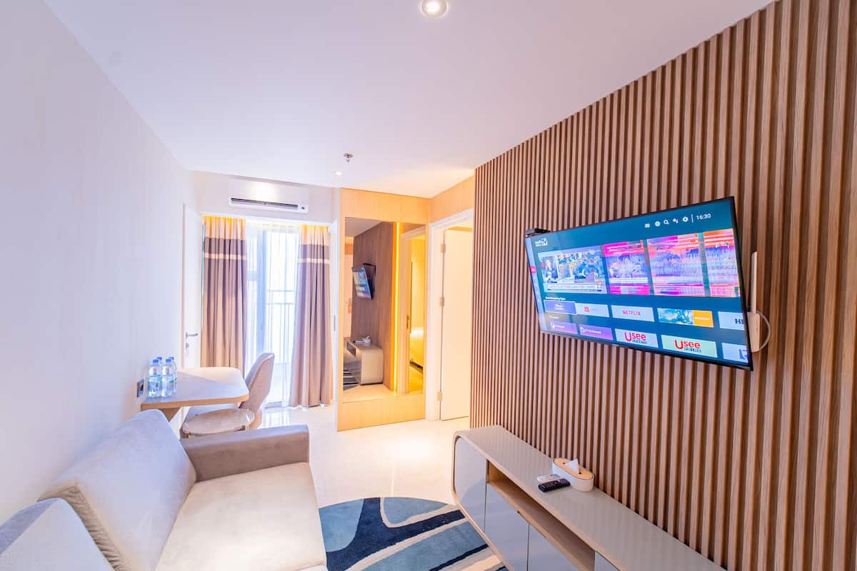 Quarto de hotel com televisão na parede exibindo diversos serviços de streaming, um sofá, uma pequena mesa com garrafas de água e uma parede revestida de madeira.