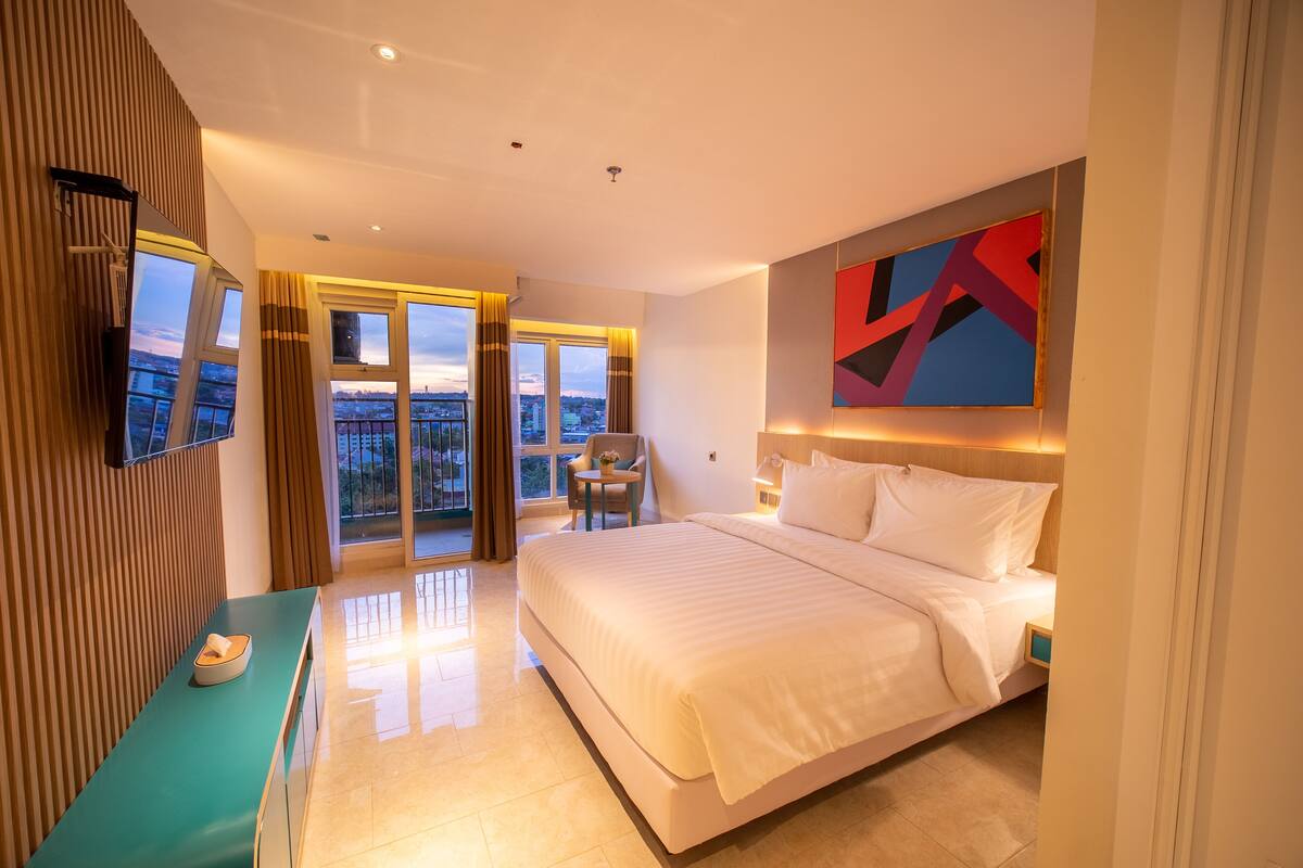 Quarto de hotel moderno com uma cama grande, televisão na parede, uma pequena mesa com uma cadeira e varanda com vista para a cidade.