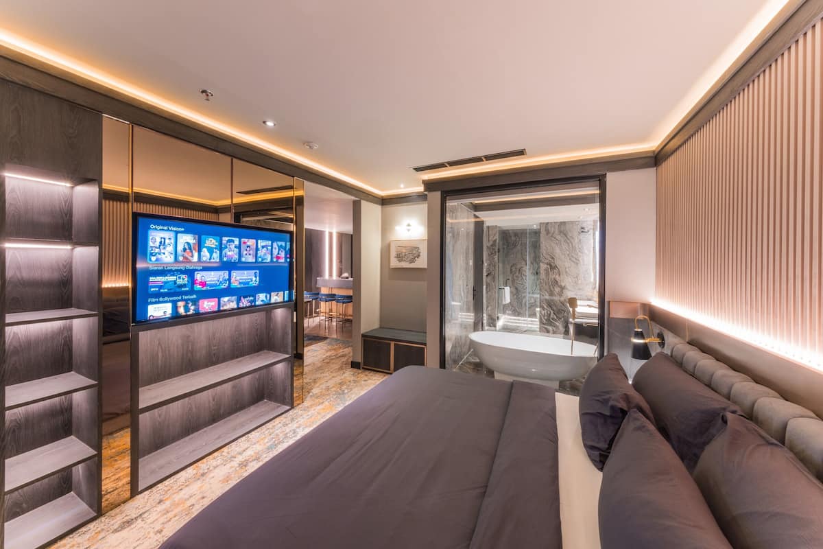 Quarto de hotel moderno com uma cama grande, uma TV exibindo um menu, um banheiro com chuveiro de vidro e uma área de estar com sofá e mesa.