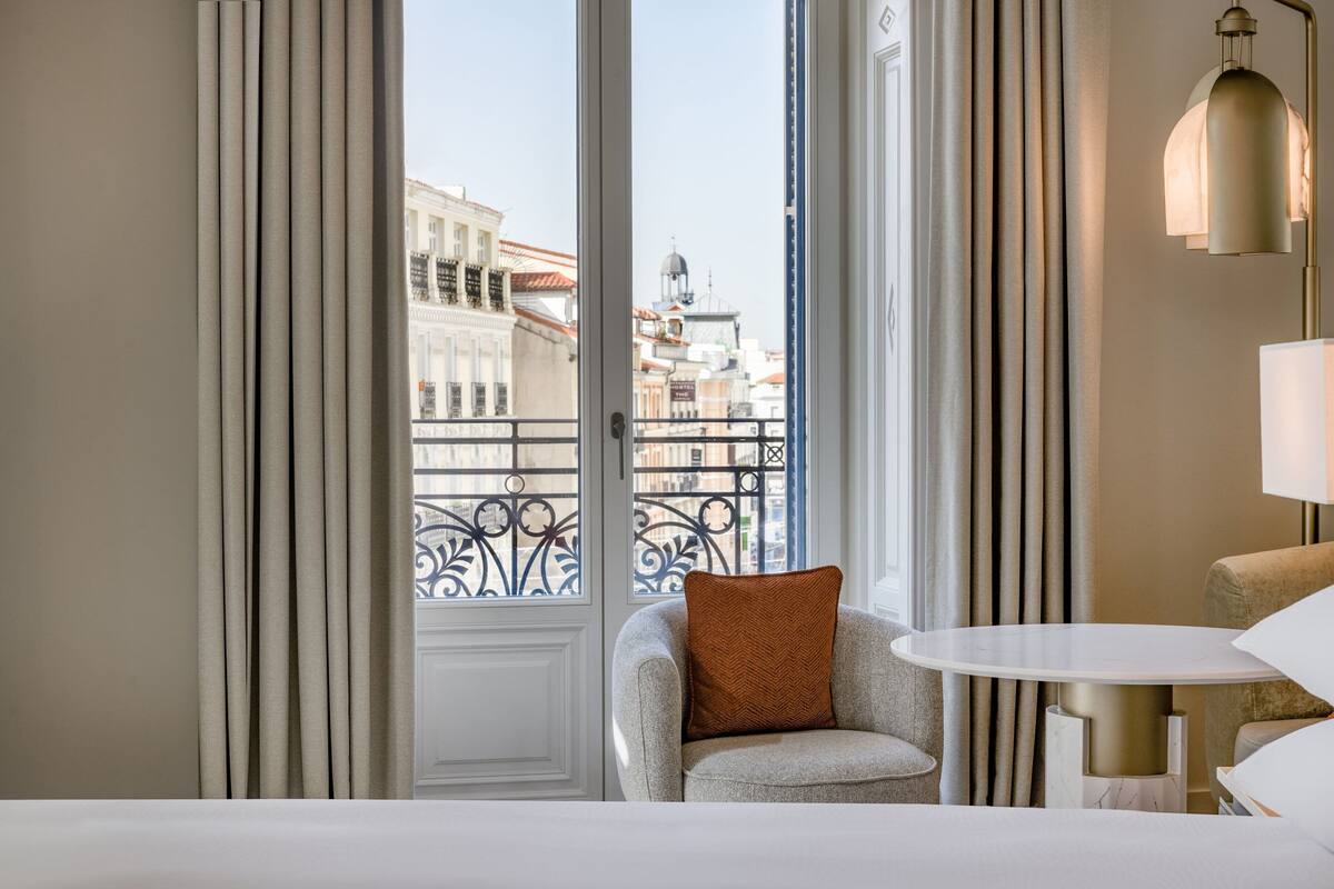 Quarto de hotel com vista para a cidade através de uma grande janela, uma mesa redonda e uma cadeira com almofada.