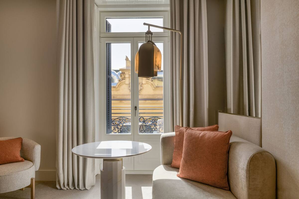 Quarto de hotel moderno com sofá, mesa redonda pequena e vista de um prédio através de uma janela grande com cortinas.