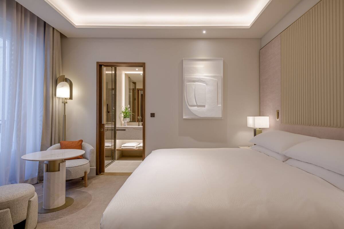 Quarto de hotel com uma cama grande, uma mesa redonda pequena, uma cadeira, um abajur, um espelho e uma porta para o banheiro.