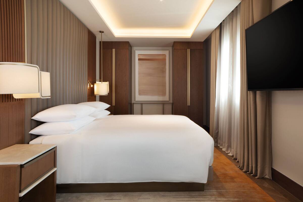 Quarto de hotel moderno com cama grande, abajures, televisão de tela plana e vista da cidade através de janelas do chão ao teto.