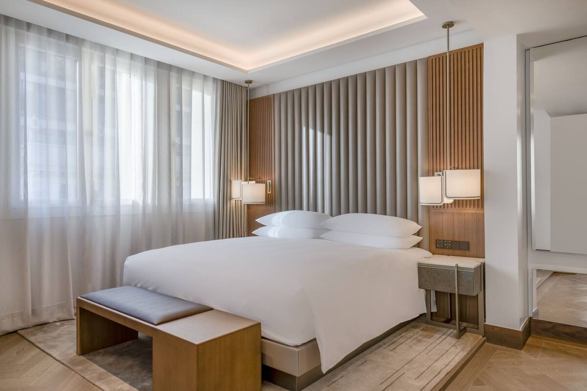 Quarto de hotel moderno com cama grande, luminárias de cabeceira, um banquinho e painel de parede de madeira.