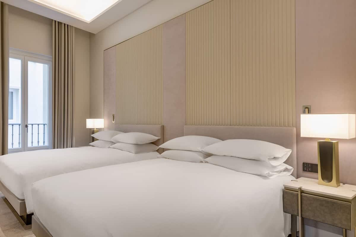 Quarto de hotel com duas camas, uma mesa de cabeceira com abajur e uma janela grande com cortinas.