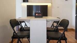 Quarto pequeno e moderno com uma mesa de madeira e quatro cadeiras pretas. Uma cesta com comida e bebida de cortesia para viajantes está colocada sobre a mesa.