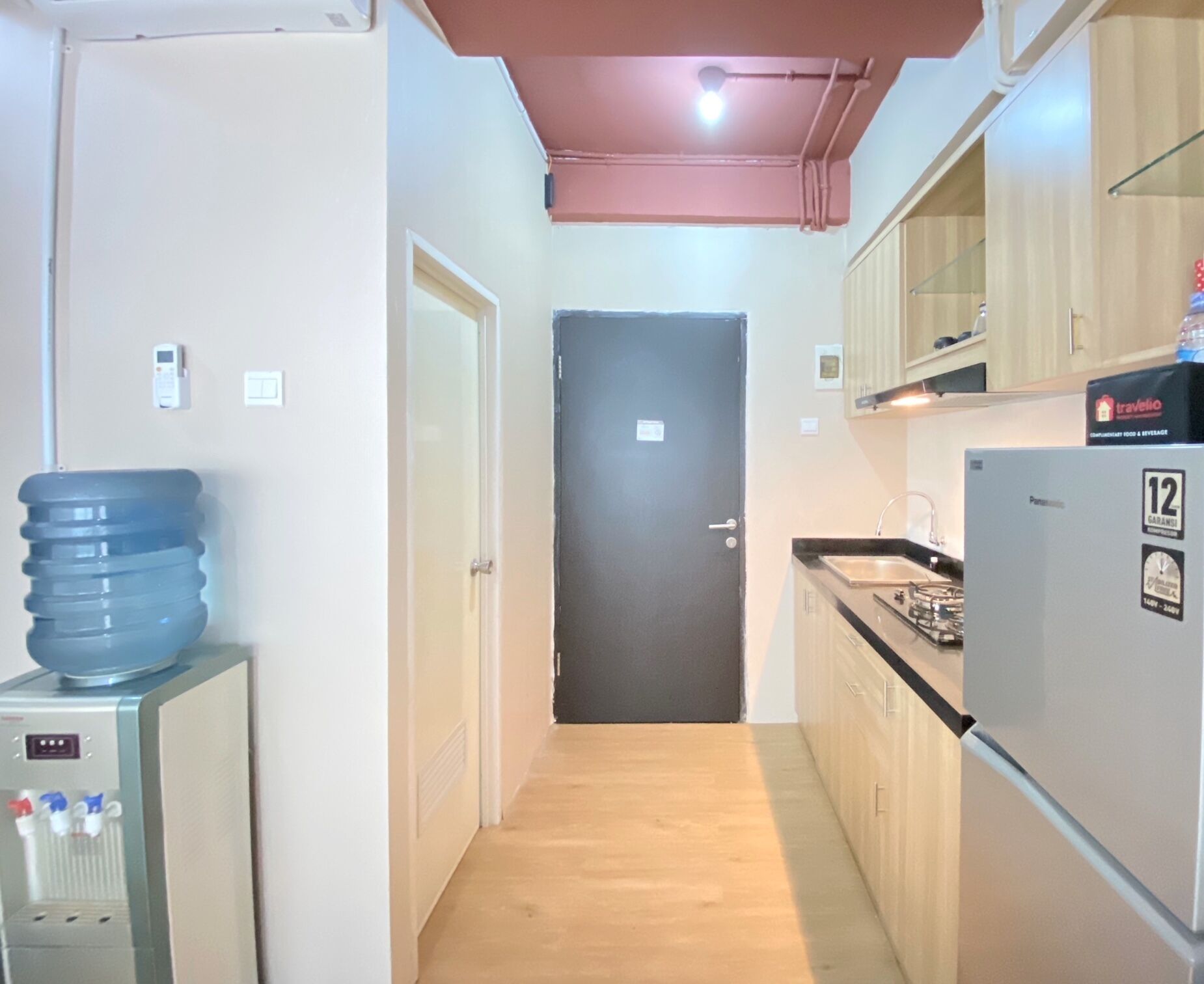 Corredor estreito com um dispensador de água à esquerda, uma porta no final e uma área de cozinha à direita.