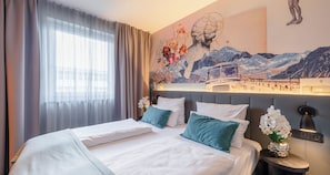 Quarto de hotel com cama, mesa de cabeceira, luminária na parede e janela com cortinas.