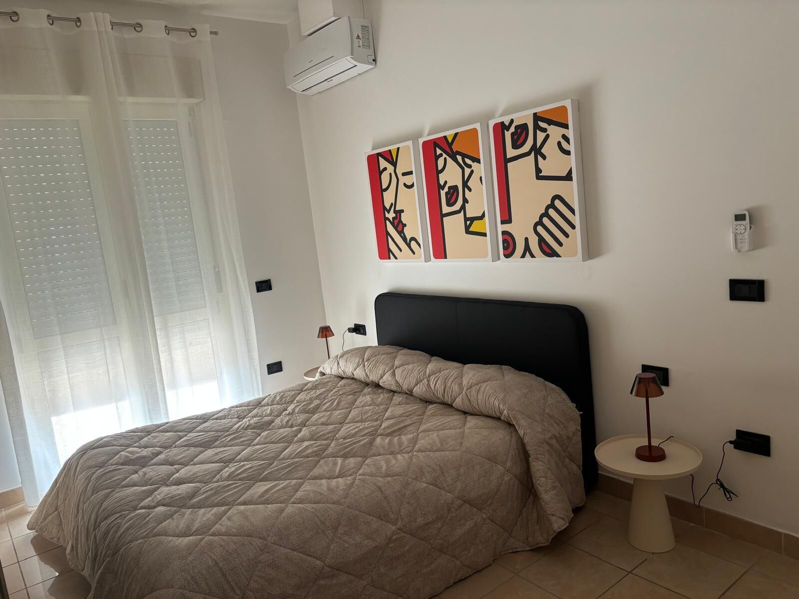 Quarto com cama, mesa de cabeceira, abajur e ar-condicionado fixado na parede.