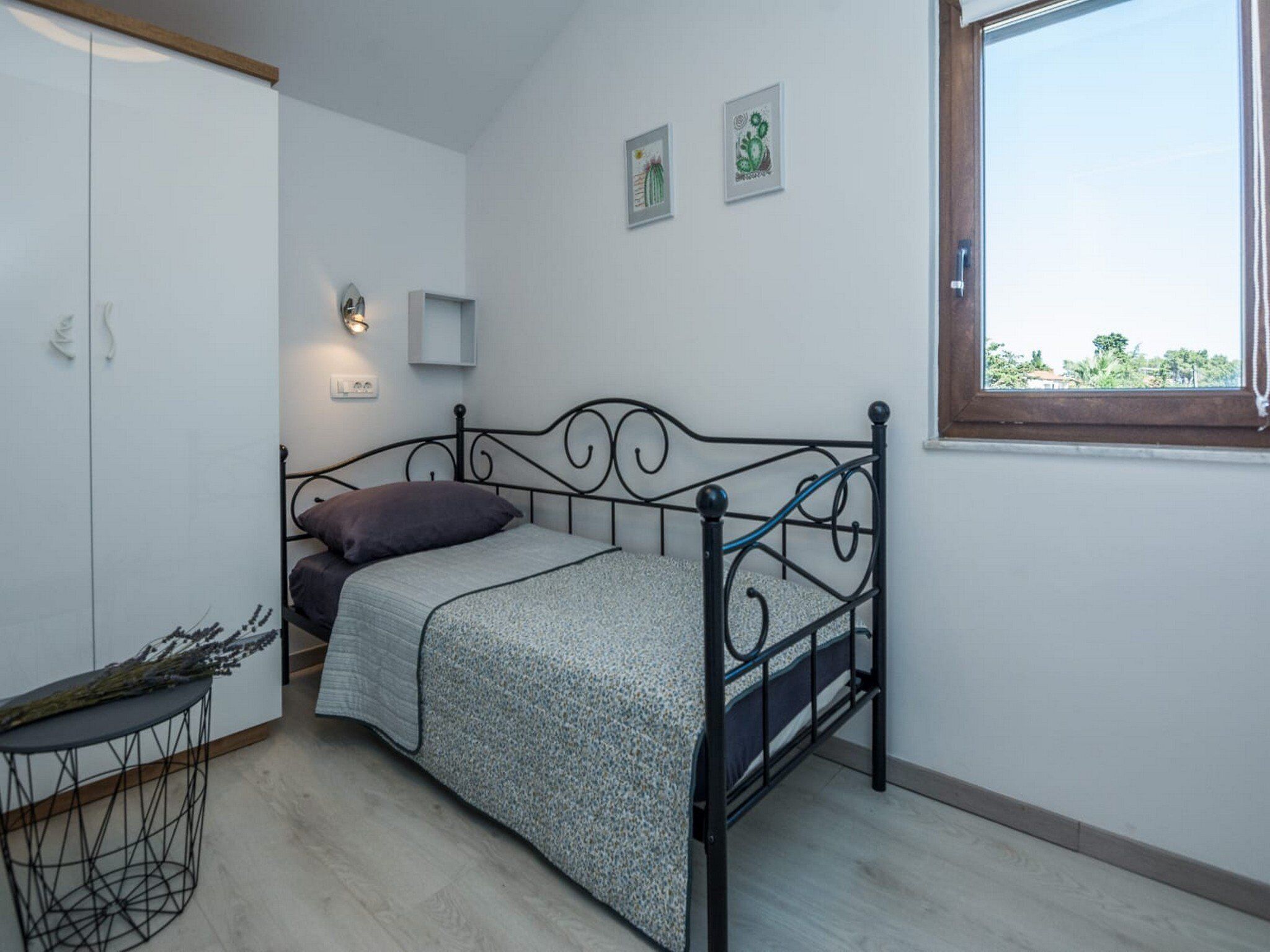 Uma cama individual com estrutura de metal, uma mesa de cabeceira, janela com vista e luminária fixada na parede.