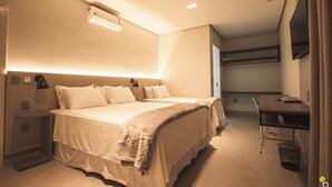 Quarto de hotel com cama grande, mesas de cabeceira, escrivaninha e TV na parede.